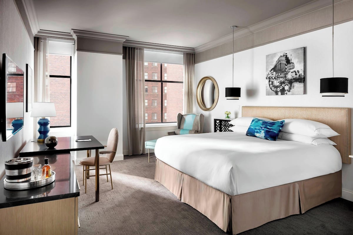 Quarto de hotel com uma cama grande, uma escrivaninha, uma cadeira, janela com cortinas e vista para a paisagem urbana.