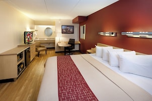 Quarto de hotel com cama, escrivaninha, cadeira, televisão, espelho e luminária na parede.