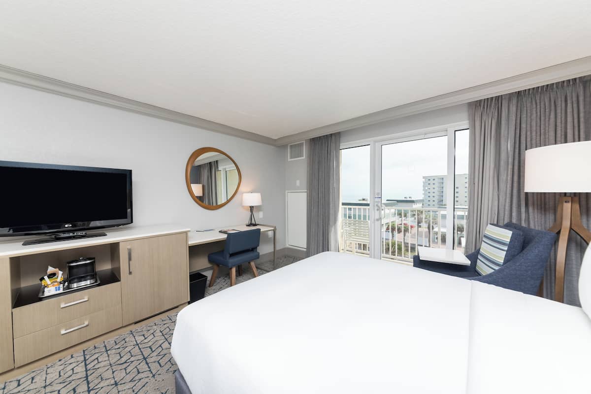 Quarto de hotel com uma cama grande, uma escrivaninha com cadeira, televisão, espelho e vista para a cidade pela janela.