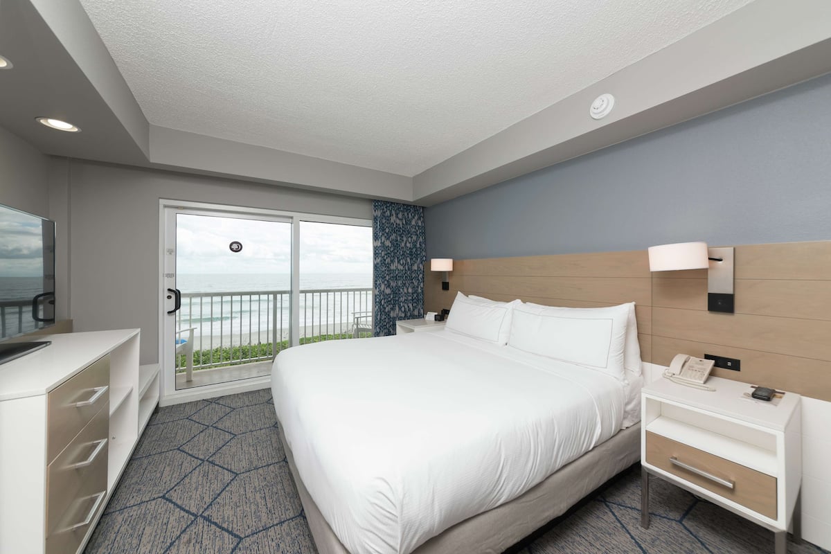 Quarto de hotel moderno com uma cama grande, uma escrivaninha com telefone e vista para o oceano por uma porta de correr.