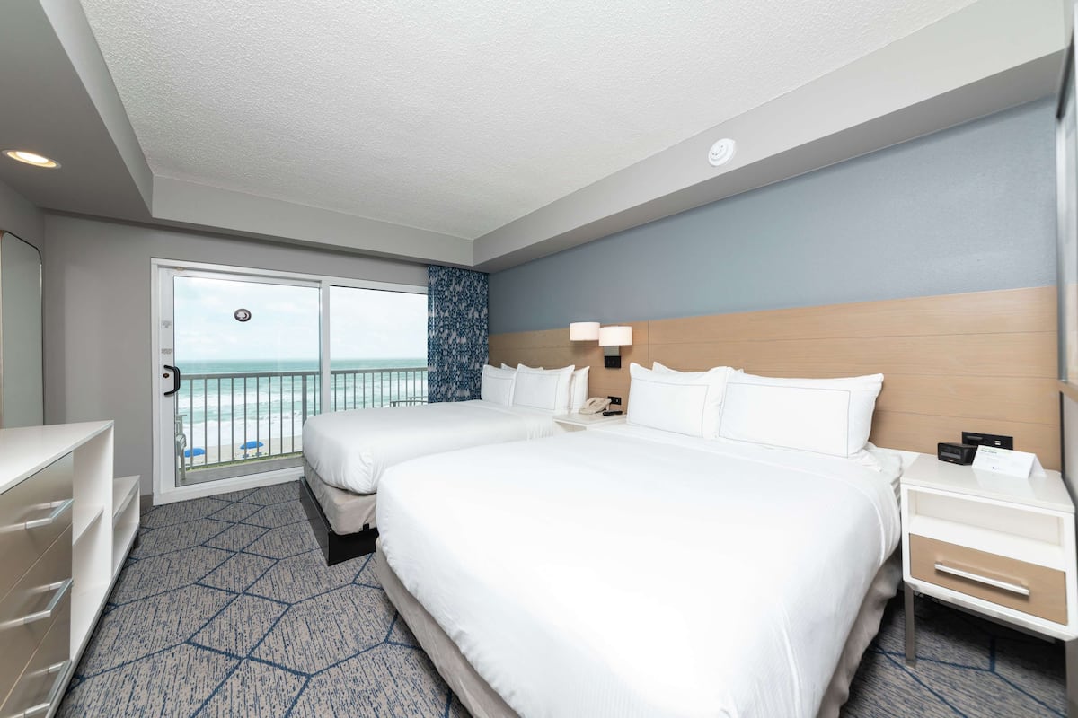 Quarto de hotel com duas camas, uma escrivaninha, cômoda e varanda com vista para o oceano.
