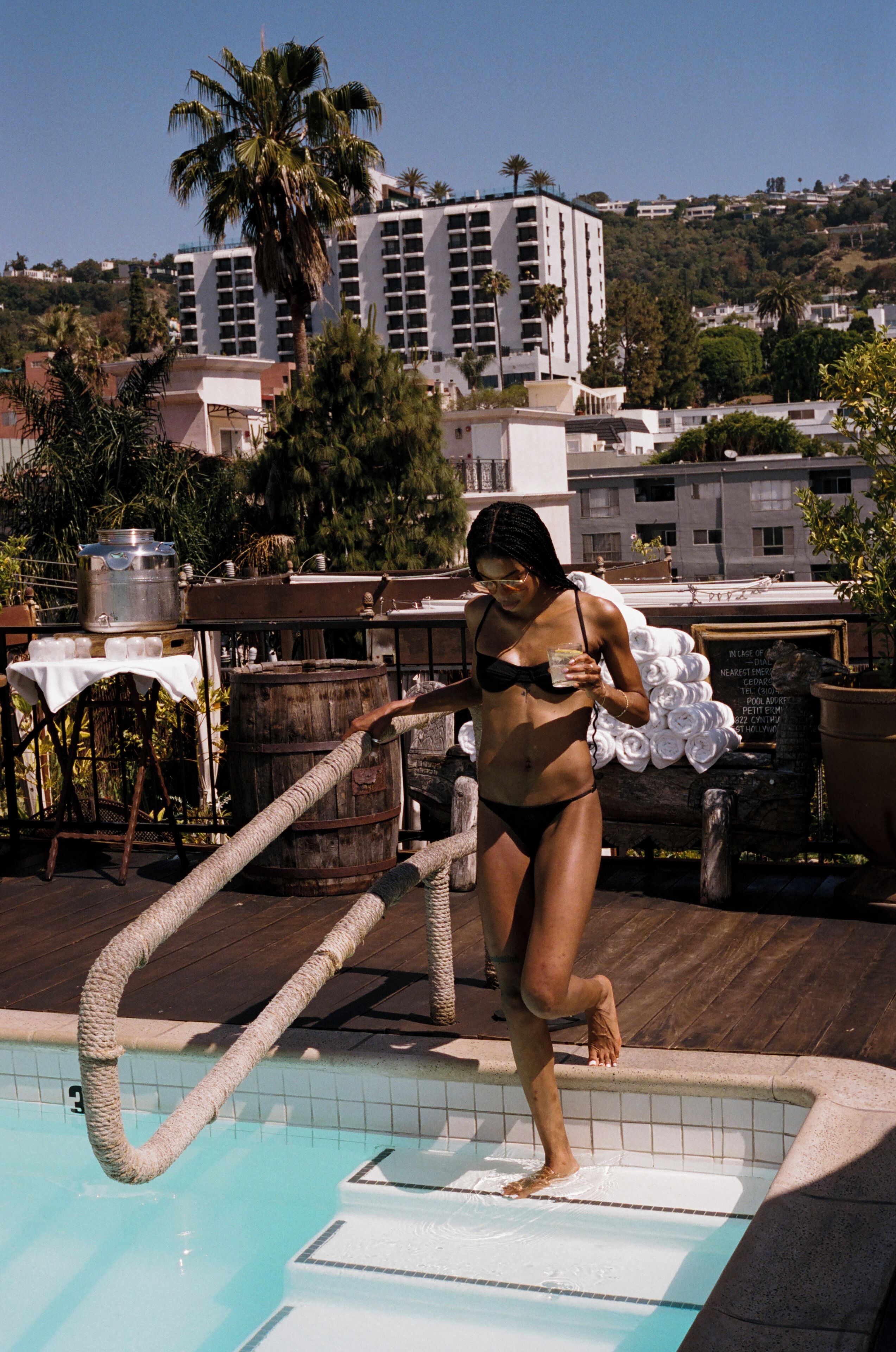 Uma mulher de biquíni preto perto de uma piscina, com um prédio branco ao fundo.