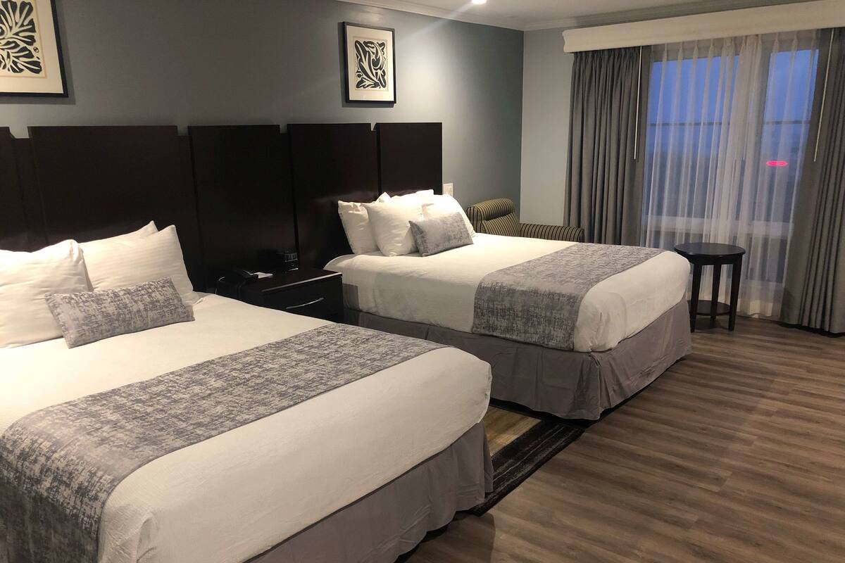 Quarto de hotel com duas camas, uma mesa de cabeceira, uma pequena mesa e uma janela grande com cortinas.