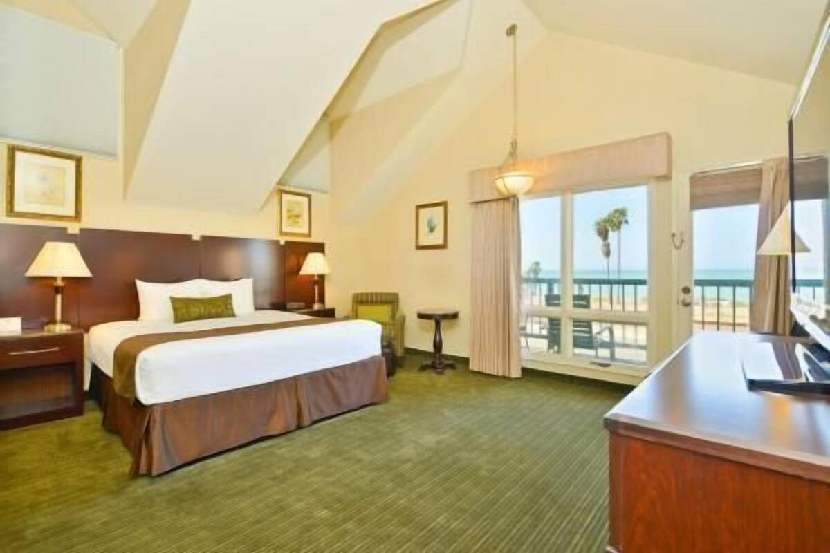 Quarto de hotel com cama, escrivaninha, cadeira, abajur, mesa de cabeceira, janela com vista para a praia e teto com claraboia.