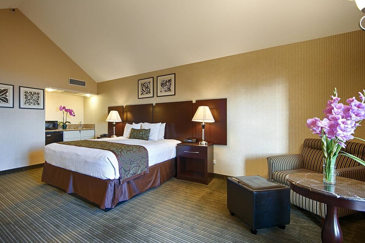 Quarto de hotel com uma cama grande, uma escrivaninha, uma cadeira, uma mesinha e um vaso de flores.