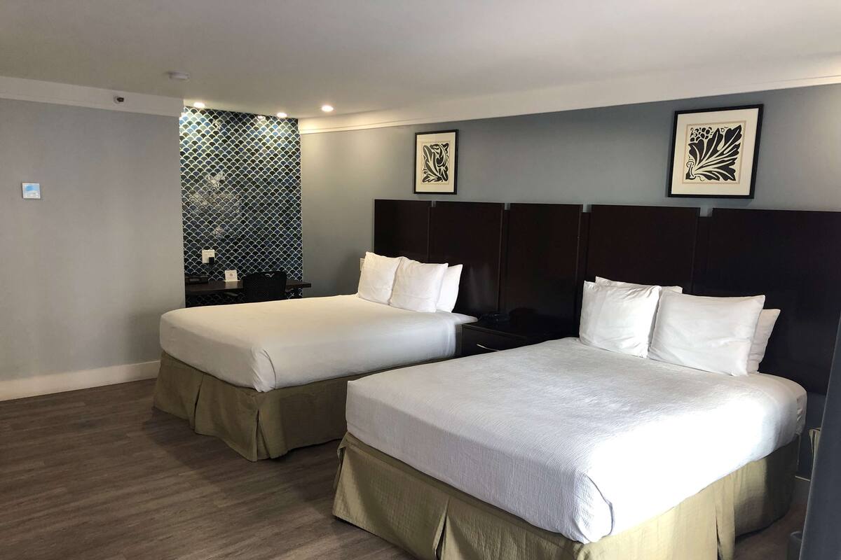 Quarto de hotel com duas camas, uma secretária pequena e um painel decorativo na parede.