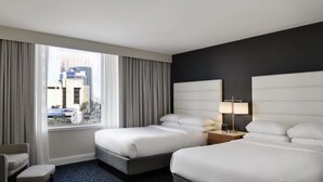 Quarto de hotel com duas camas, uma cadeira, uma mesinha e vista para a cidade através de janelas amplas.