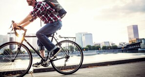 Uma pessoa andando de bicicleta com uma mochila, um horizonte urbano ao fundo e um corpo de água.