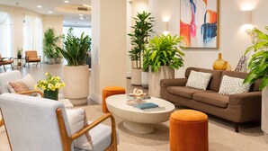 Sala de estar moderna com sofá, poltrona, mesa de centro e pufes laranja. Há plantas em vasos e um quadro na parede.