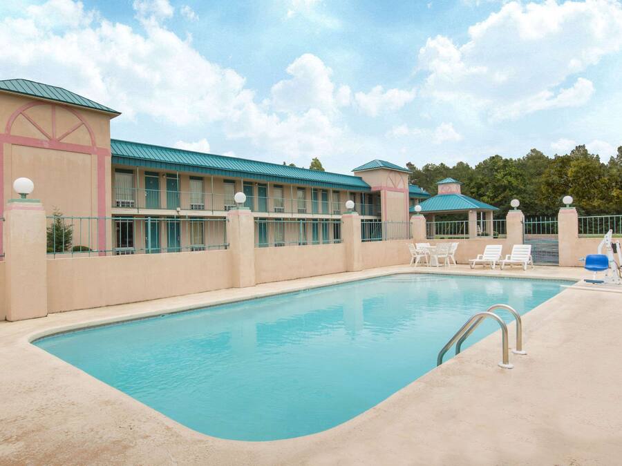 Piscina com área ao redor, espreguiçadeiras e um edifício com telhado verde e paredes bege.