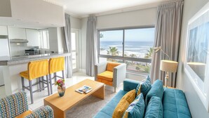 Sala de estar moderna com uma janela grande, cozinha ao fundo e vista para o mar.