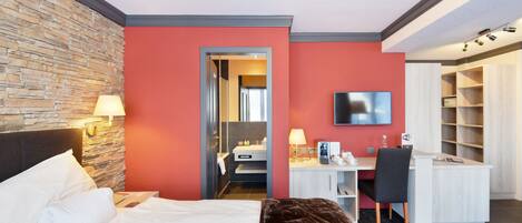 Quarto de hotel com cama, escrivaninha, cadeira, televisão e guarda-roupa.