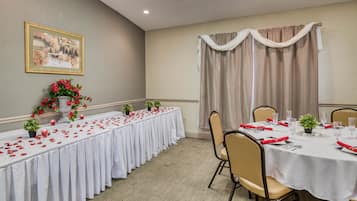 Uma sala de conferências com uma mesa comprida, cadeiras e toalha de mesa branca. Há um quadro na parede e uma janela com cortinas.