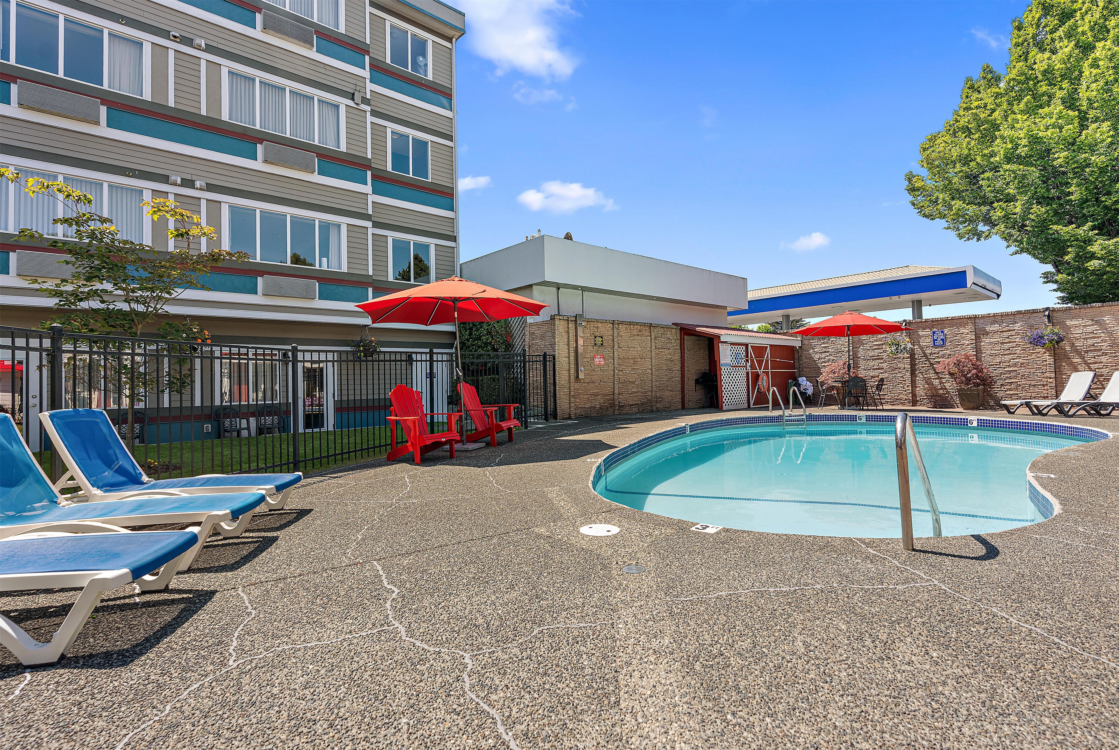 Área de piscina de hotel com espreguiçadeiras, um guarda-sol vermelho e um edifício com várias janelas.