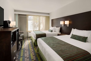 Quarto de hotel com duas camas, uma escrivaninha, uma cadeira, uma televisão e uma janela com cortinas.