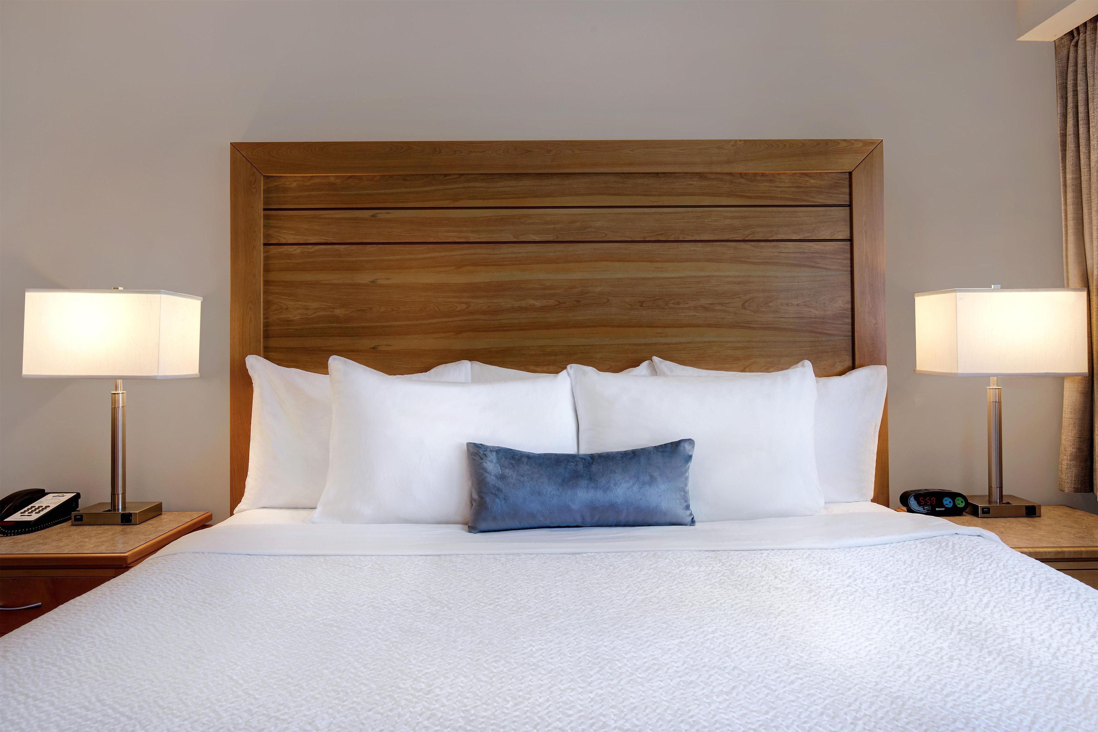 Quarto de hotel com uma cama grande, duas luminárias laterais, telefone ao lado da cama e relógio digital.