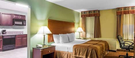 Quarto de hotel com cama, luminárias de cabeceira, escrivaninha com telefone, cadeira e cozinha compacta com micro-ondas.