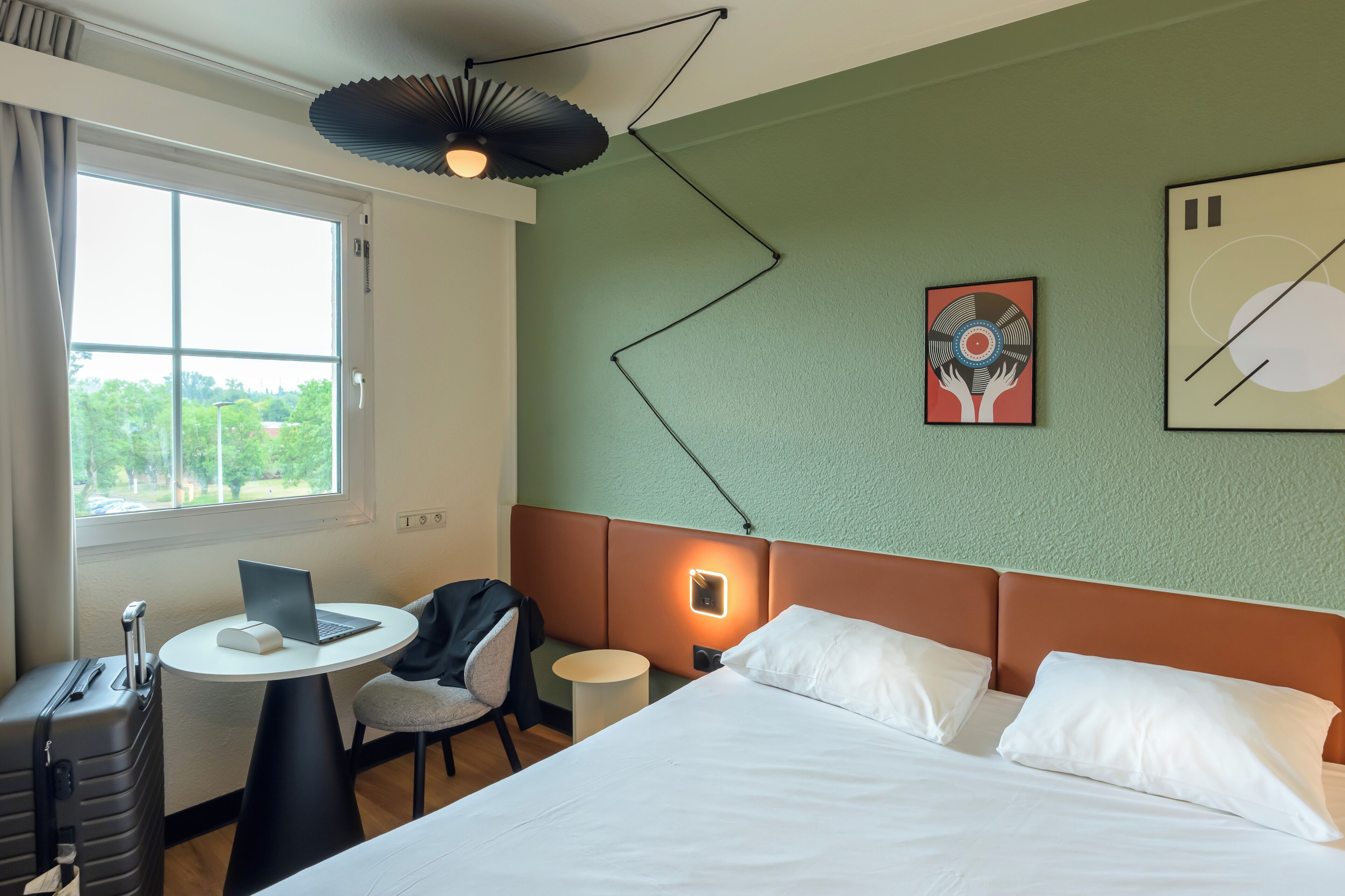 Quarto de hotel com cama, secretária com um laptop, cadeira, mala e obras de arte nas paredes.