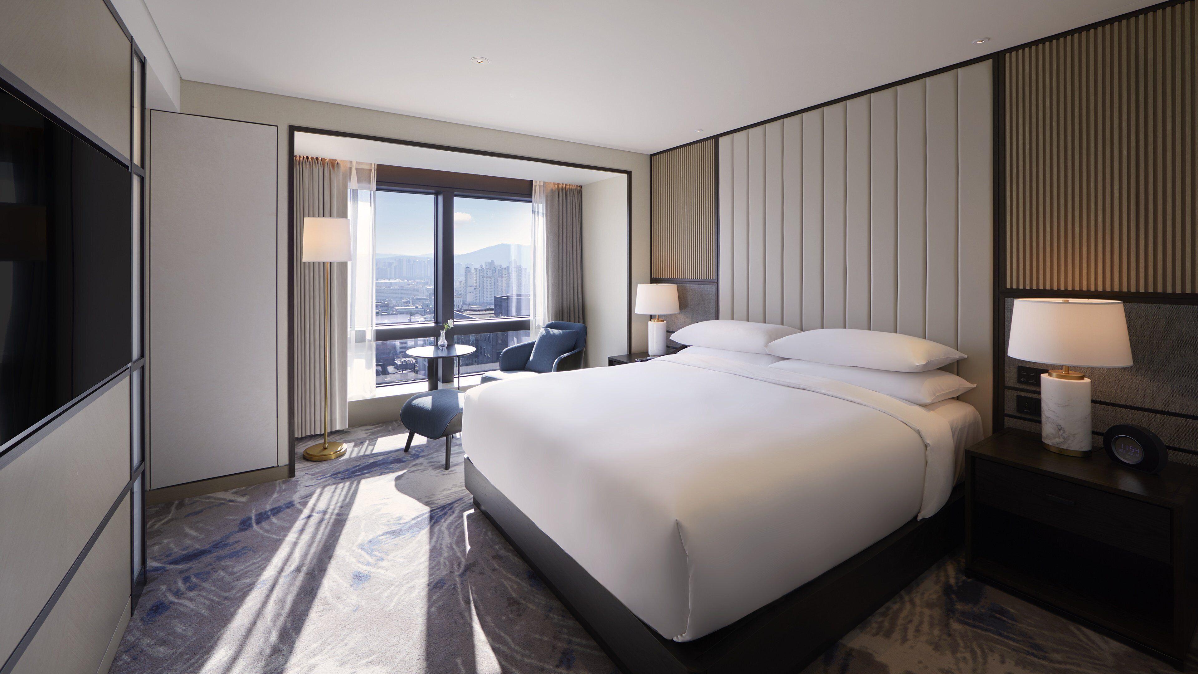 Quarto de hotel moderno com uma cama grande, duas mesas de cabeceira, uma televisão, janela com cortinas e vista para a cidade.