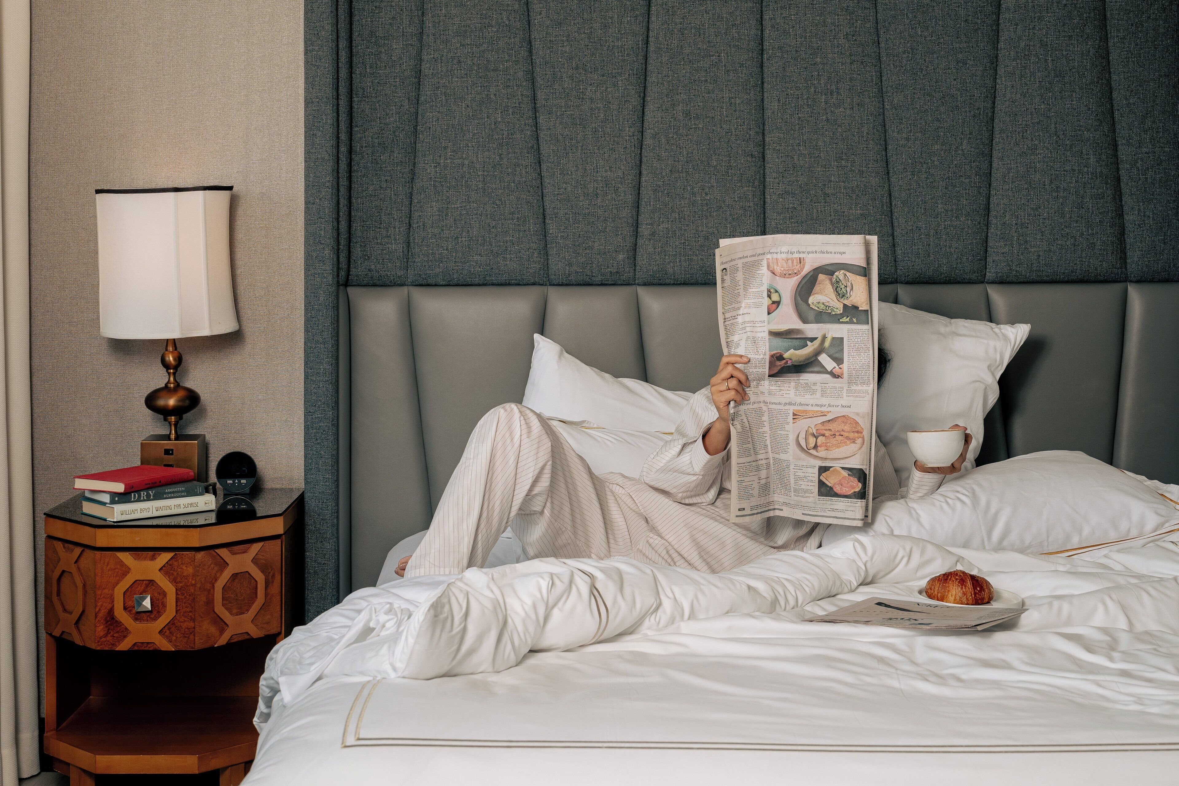 Quarto de hotel com uma cama, uma mesa de cabeceira com abajur, um jornal e um croissant sobre a cama.