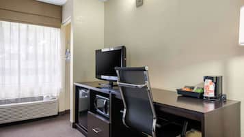 Quarto de hotel com escrivaninha, televisão, cadeira, cafeteira e janela com cortinas.