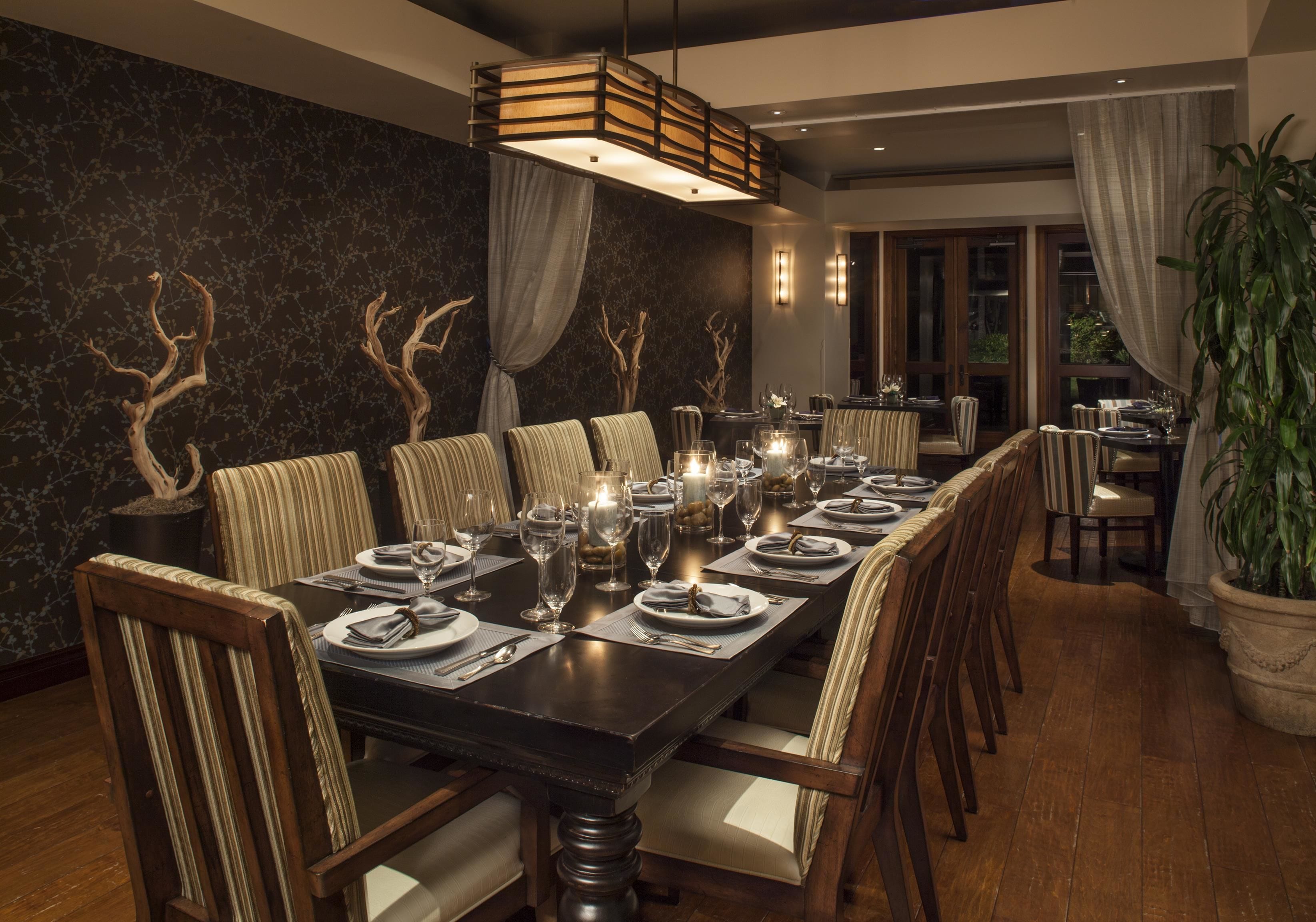 Uma sala de jantar com uma mesa comprida posta para refeição, rodeada por cadeiras estofadas.