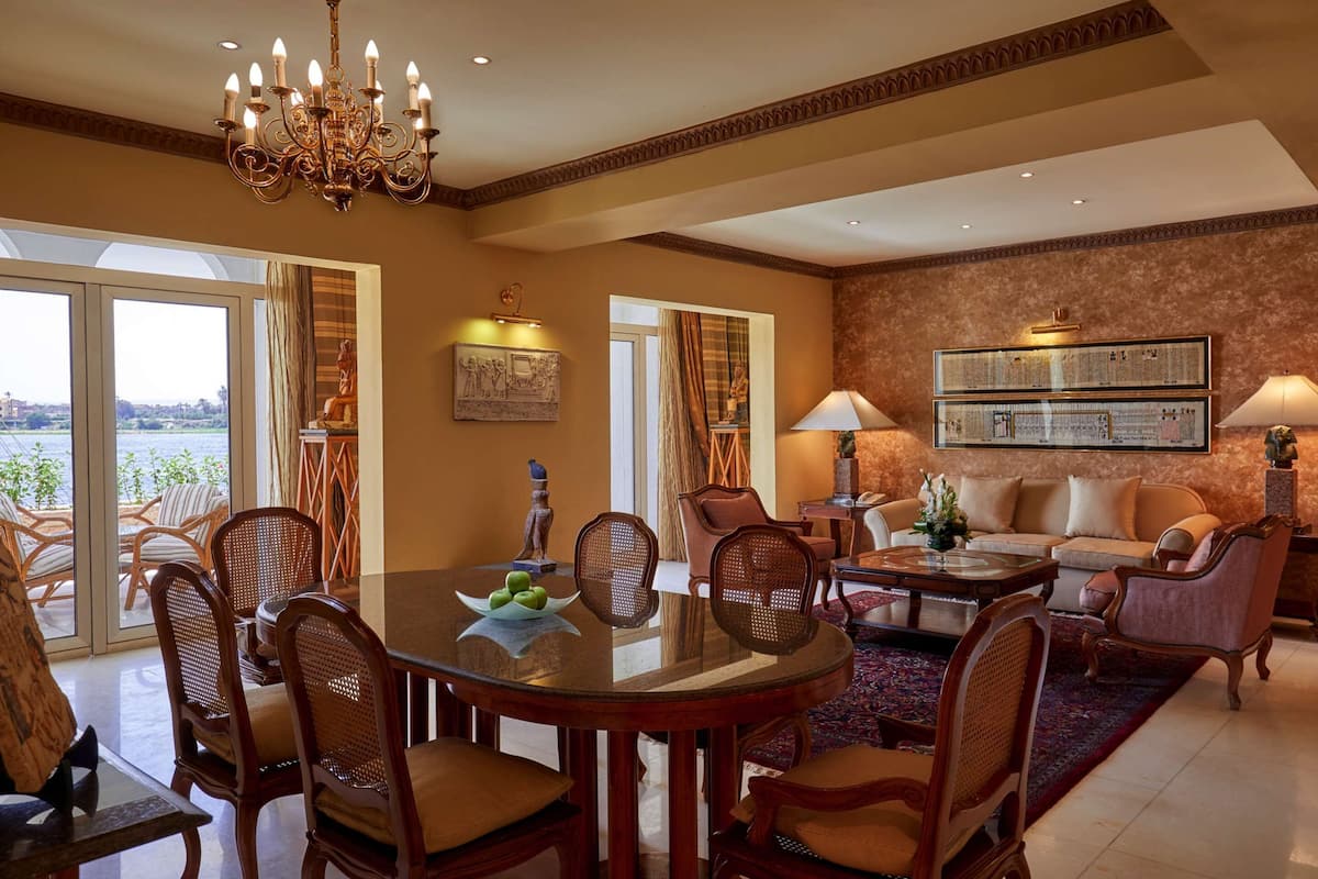 Uma sala de jantar com uma mesa redonda, cadeiras e um candelabro. Há um sofá e um abajur na sala de estar.