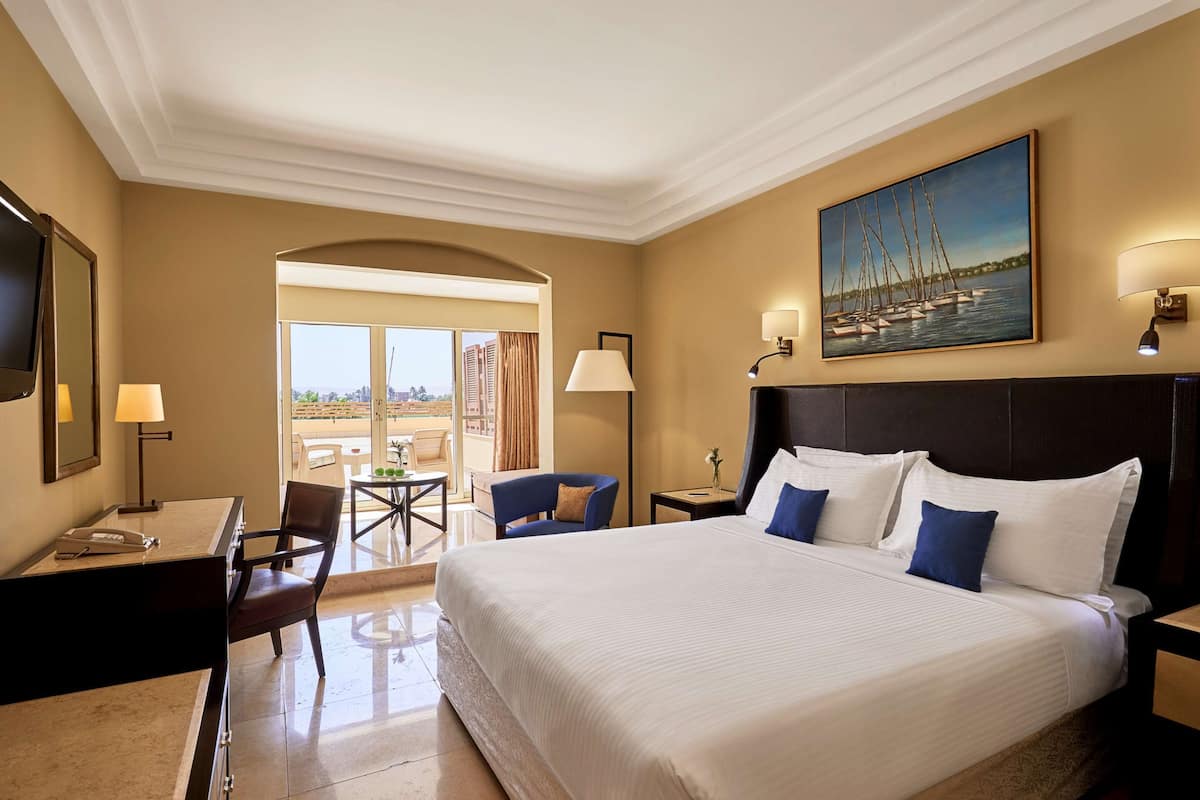Quarto de hotel com uma cama grande, uma escrivaninha com telefone, uma cadeira, uma mesa e um quadro de barcos à vela na parede.