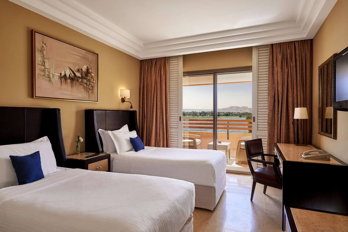 Quarto de hotel com duas camas, uma escrivaninha, uma cadeira, luminária de mesa, mesa de cabeceira, uma obra de arte emoldurada e uma janela grande com cortinas.
