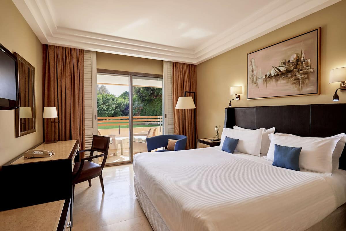Quarto de hotel com uma cama grande, uma escrivaninha com telefone, uma cadeira, uma mesa, um abajur e um quadro na parede.
