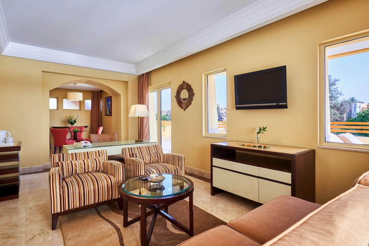 Quarto de hotel com sofá, poltronas, mesa de centro de vidro, televisão, escrivaninha com luminária e vista para o exterior.