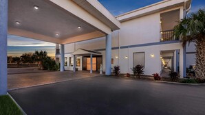 Um hotel moderno com entrada coberta, varanda no segundo andar e um acesso arborizado.