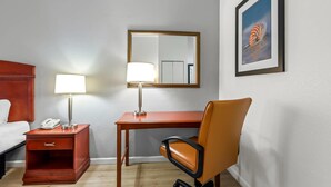 Quarto de hotel com cama, escrivaninha com luminária, cadeira, mesa de cabeceira com telefone, espelho e quadro na parede.