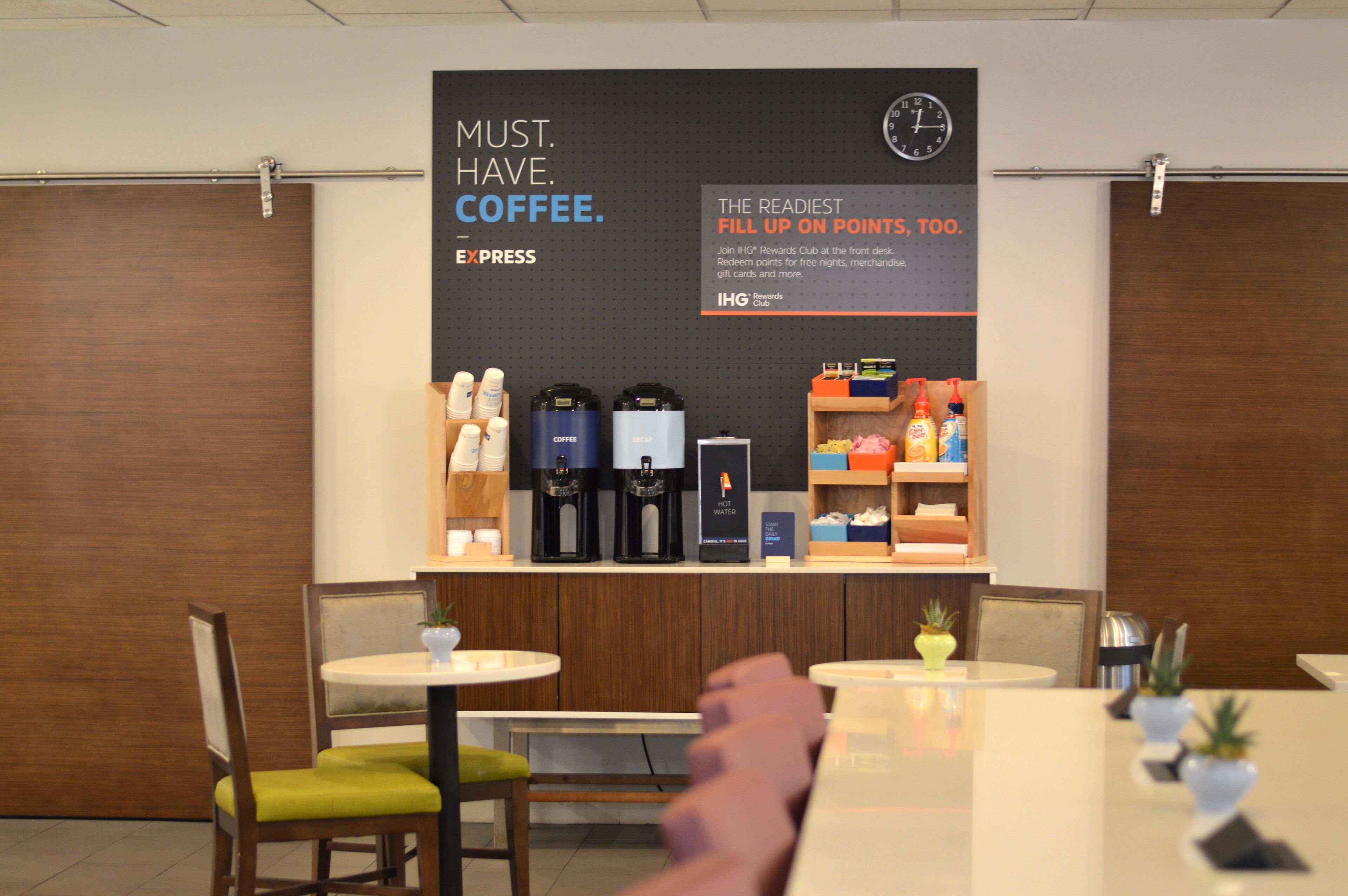 Uma estação de café com um aviso divulgando um programa de recompensas, um relógio e diversos dispensadores de café e água.