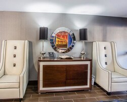 Um hall de hotel com dois poltronas bege, uma mesa consola de madeira e um relógio fixado na parede.