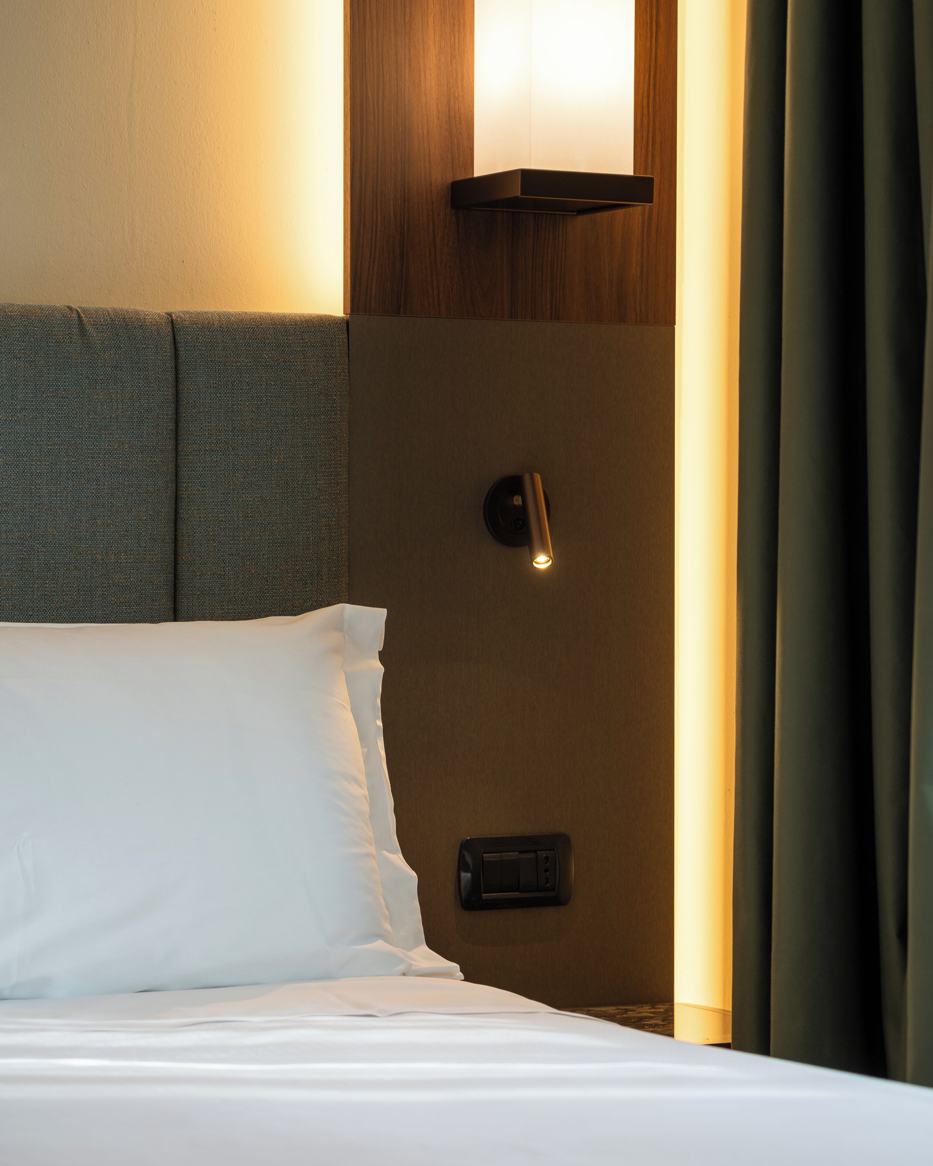 Quarto de hotel com uma cama bem arrumada, luminária fixada na parede e painel de controle.