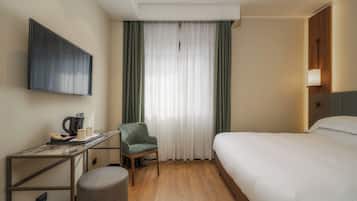Quarto de hotel com uma cama, uma mesa com cafeteira, uma cadeira e uma televisão na parede.