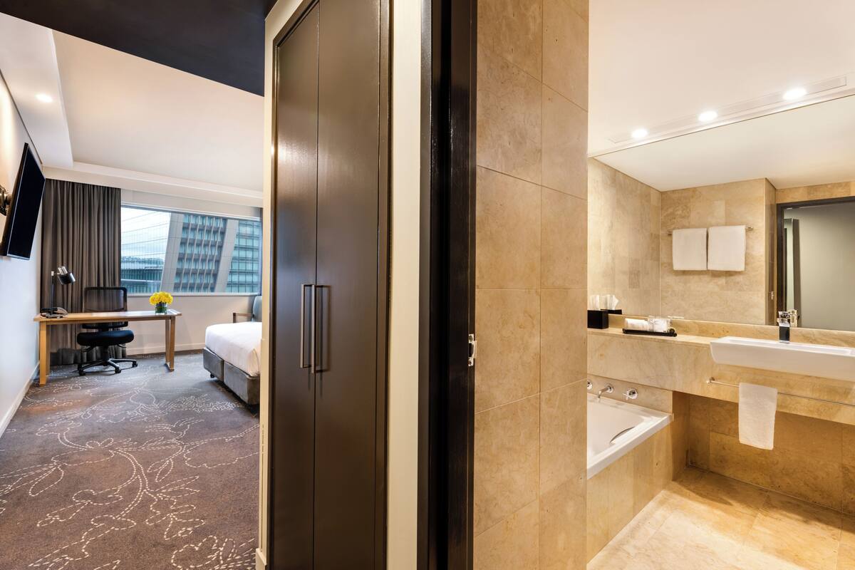 Quarto de hotel com cama, escrivaninha, cadeira e vista para um edifício.