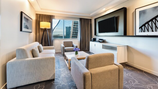 Quarto de hotel com sofá, poltronas, mesa de centro, televisão de tela plana, escrivaninha e uma grande janela com cortinas.