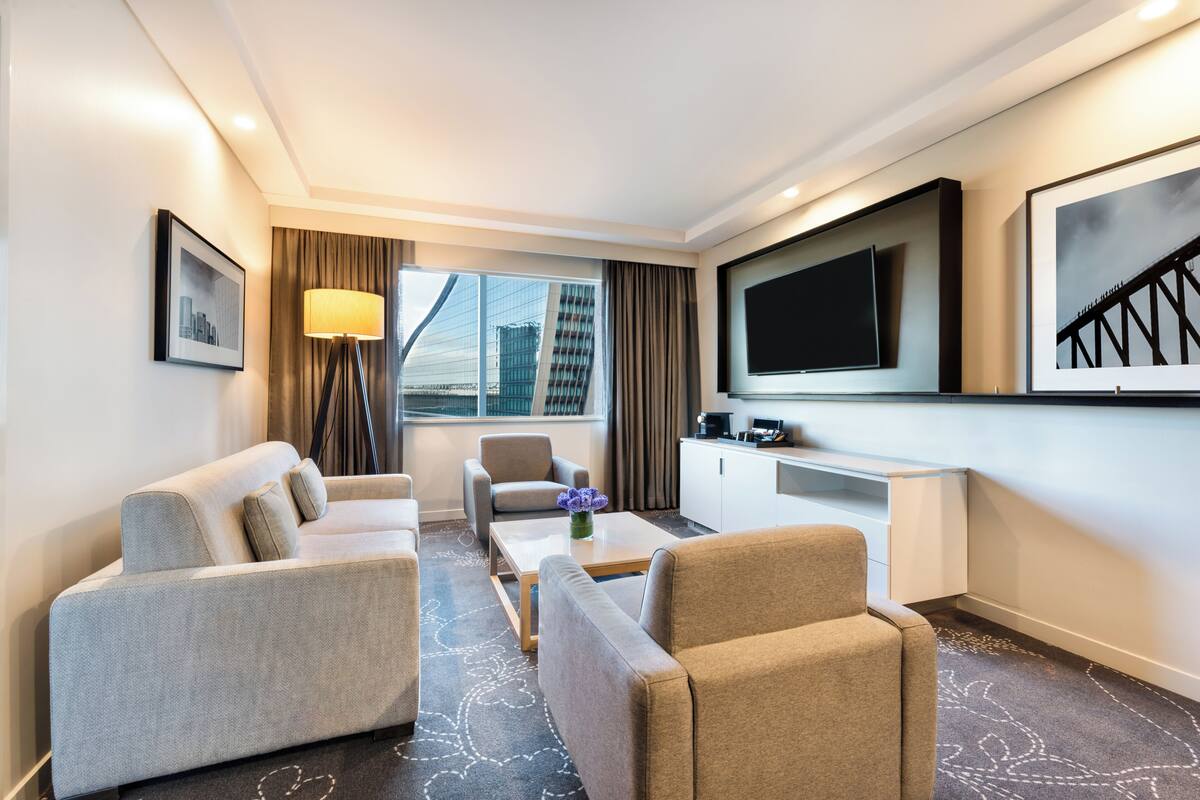 Quarto de hotel com sofá, poltronas, mesa de centro, televisão de tela plana, escrivaninha e uma grande janela com cortinas.