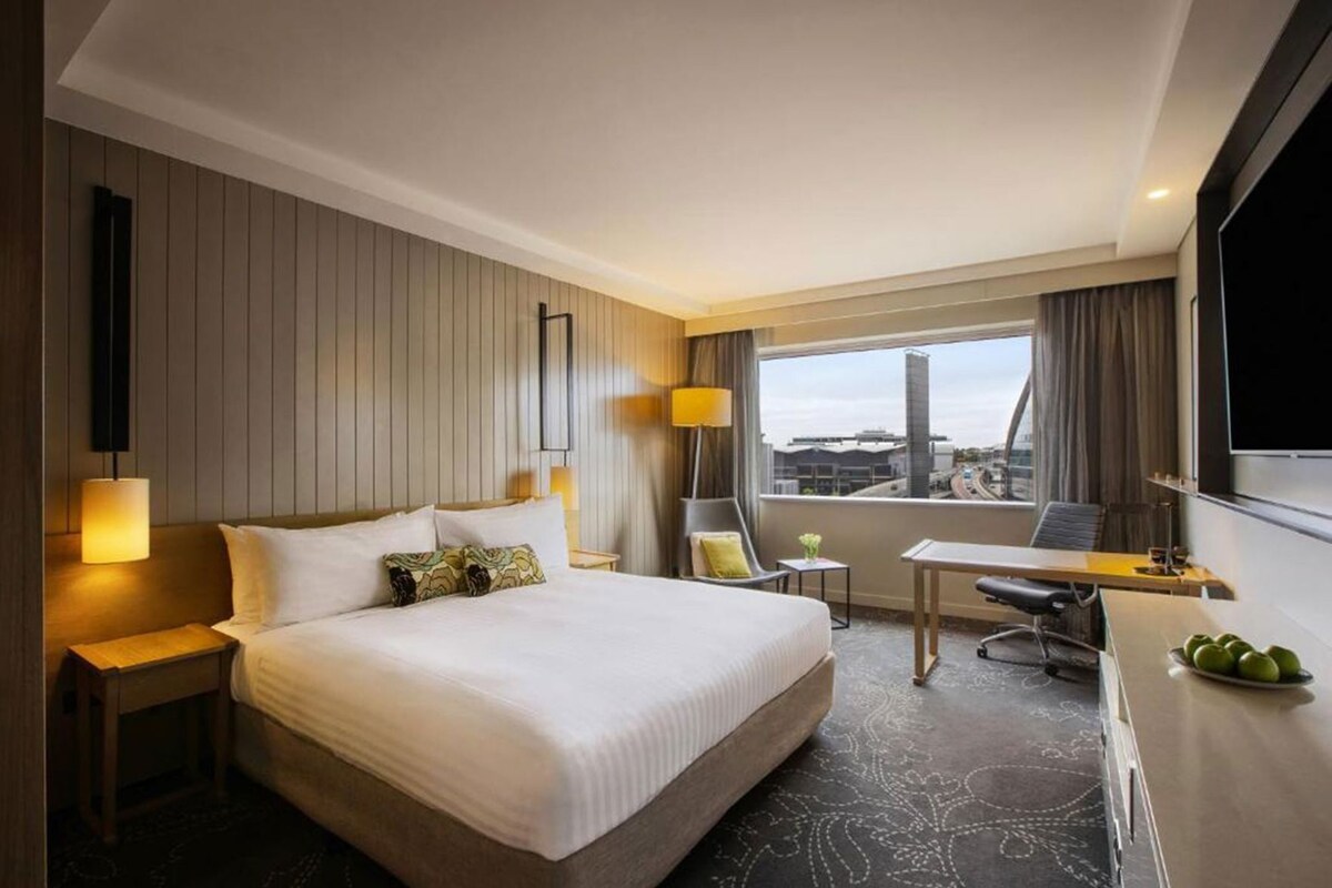 Quarto de hotel com uma cama grande, uma escrivaninha, uma cadeira, um abajur e vista para a paisagem urbana.