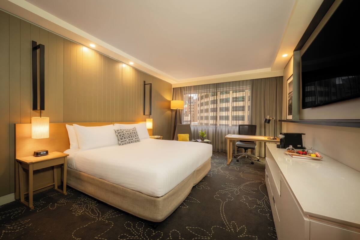 Quarto de hotel com uma cama grande, mesas de cabeceira com luminárias, uma escrivaninha, televisão e uma pequena área de cozinha.