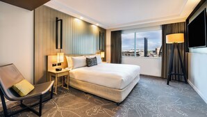 Quarto de hotel com cama, poltrona, mesa de cabeceira, luminária, televisão e vista da cidade.