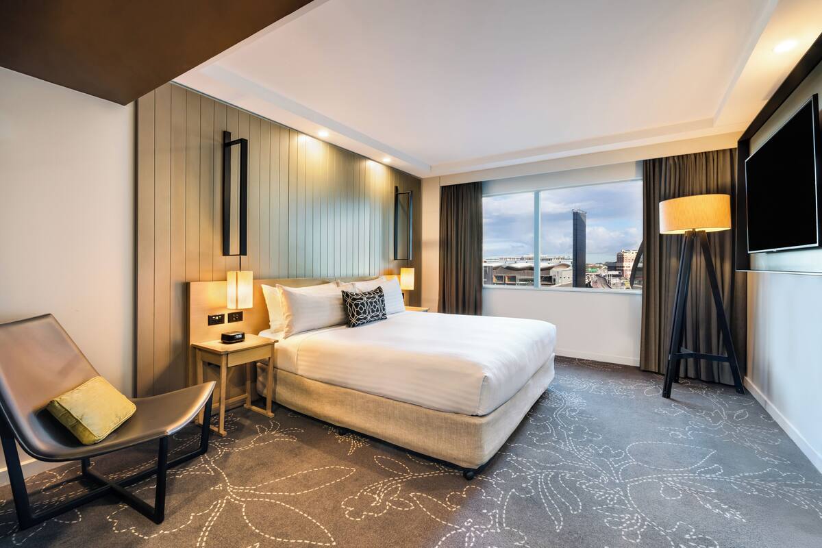 Quarto de hotel com cama, poltrona, mesa de cabeceira, luminária, televisão e vista da cidade.