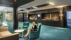 Um hall de hotel moderno com carpete verde-água, um espaço de estar com uma mesa redonda e um grande pilar.