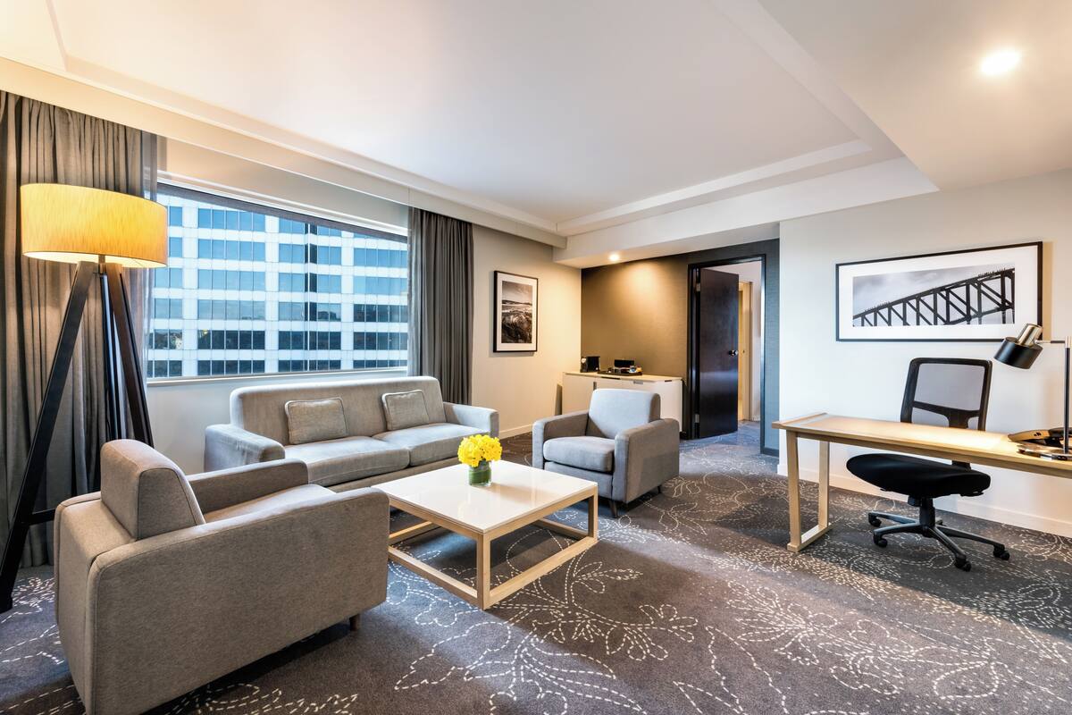 Quarto de hotel com sofá, mesa de centro, escrivaninha, cadeira, luminária, porta-retrato e vista de um edifício pela janela.