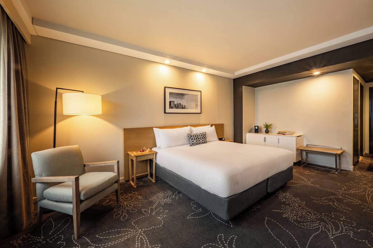 Quarto de hotel com uma cama grande, uma cadeira, uma mesa de cabeceira, um abajur, um quadro na parede e um tapete com padrão floral.