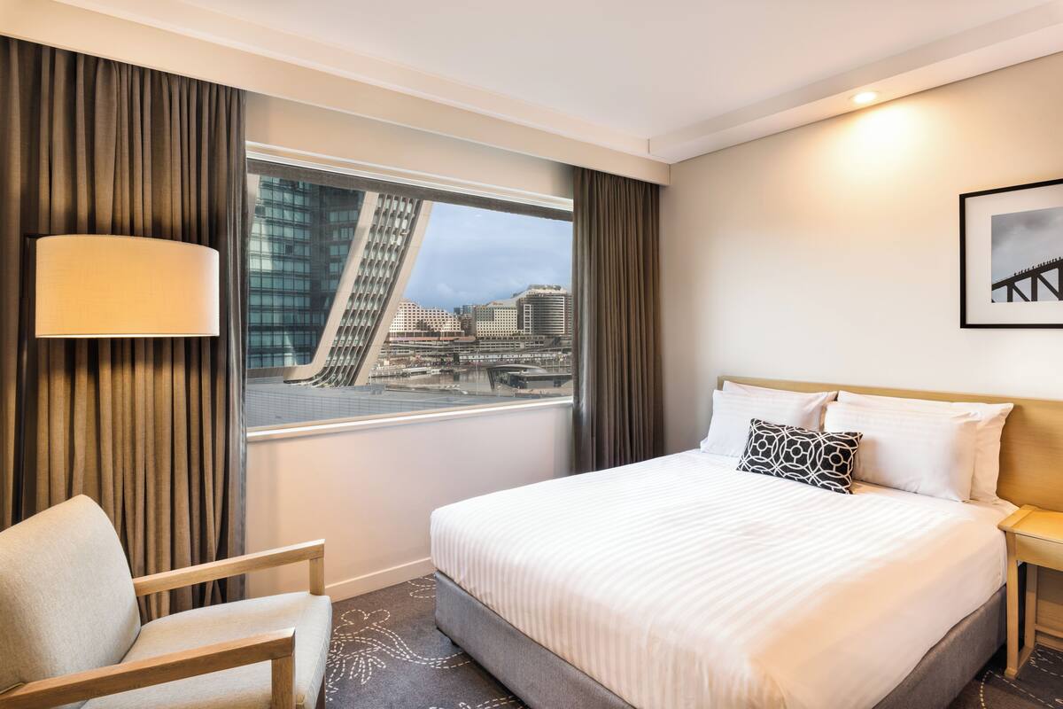 Quarto de hotel com uma cama grande, uma cadeira, uma janela com vista para a cidade e um abajur.