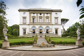 Um edifício amplo e simétrico, com uma escadaria central, rodeado por um jardim bem cuidado e estátuas.