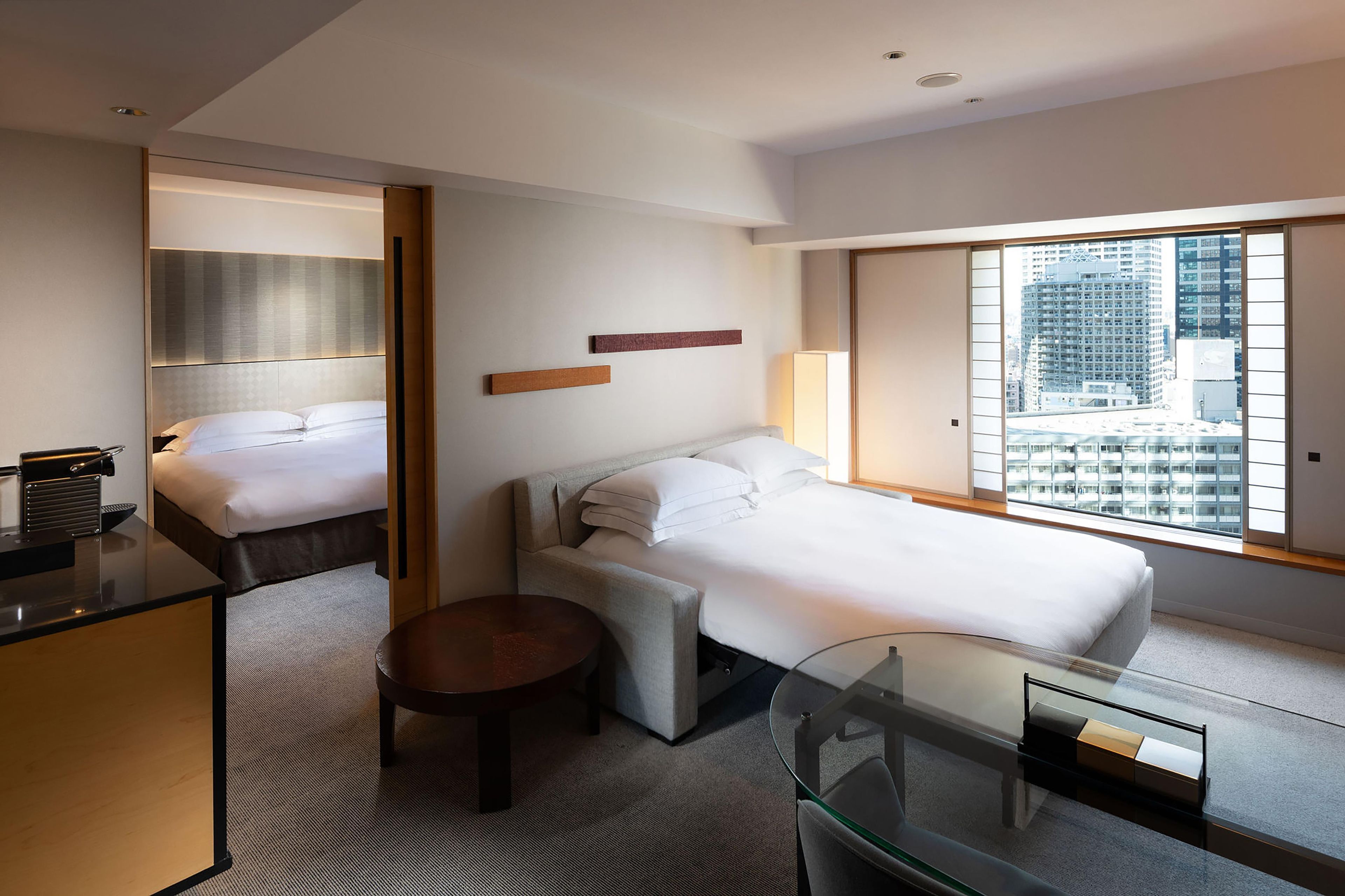 Quarto de hotel com cama, escrivaninha, cadeira, mesinha de centro e janela com vista para a cidade.